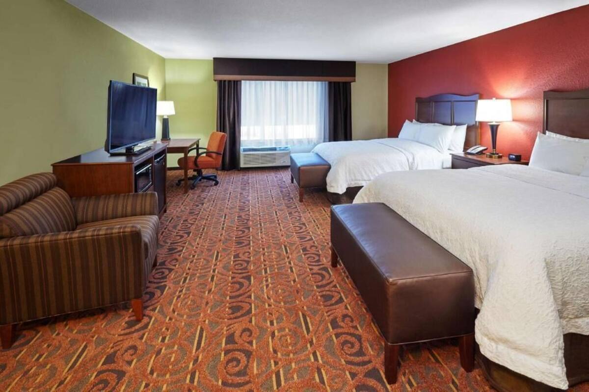 Quarto de hotel com uma cama grande, um sofá, uma mesa de trabalho com uma televisão e uma janela com cortinas.