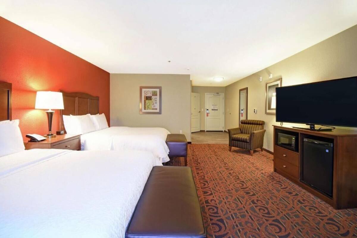 Quarto de hotel com duas camas, uma escrivaninha, uma cadeira, uma televisão e piso coberto por carpete.