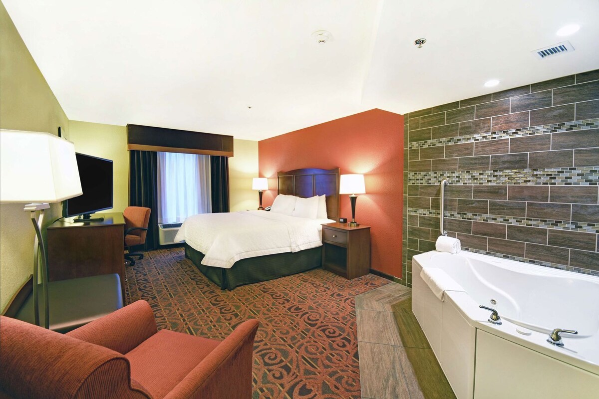 Quarto de hotel com cama, escrivaninha com computador, cadeira, abajur, mesa de cabeceira, banheiro com banheira e chuveiro.