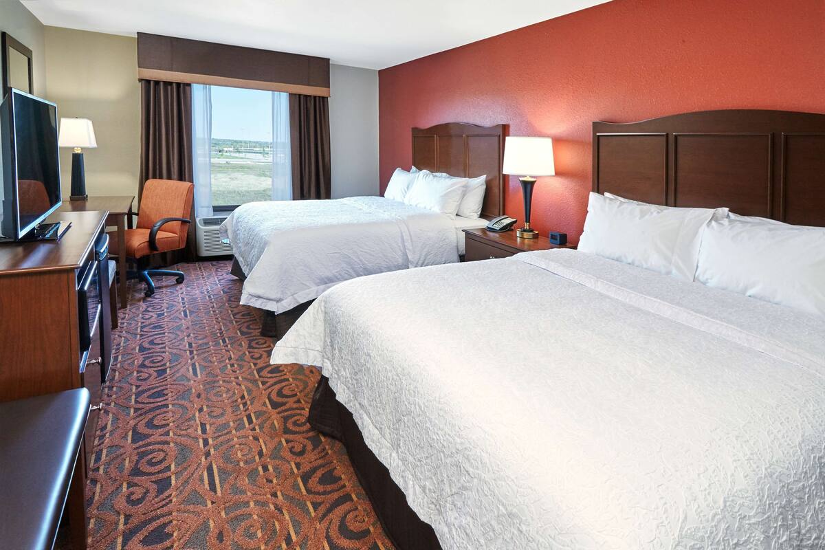 Quarto de hotel com duas camas, uma escrivaninha com televisão, uma cadeira e piso coberto por carpete.