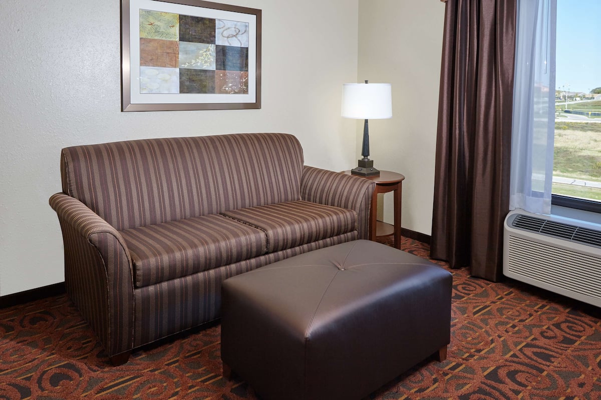 Quarto de hotel com um sofá listrado, uma mesa pequena com uma luminária, um tapete estampado e uma janela com cortinas.