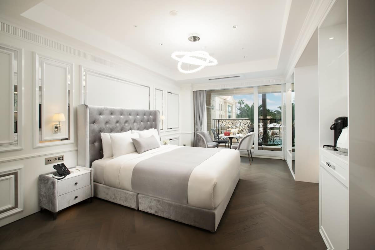 Quarto de hotel moderno com uma cama grande, mesa de cabeceira com telefone, varanda com mesa e cadeiras e vista para o exterior.