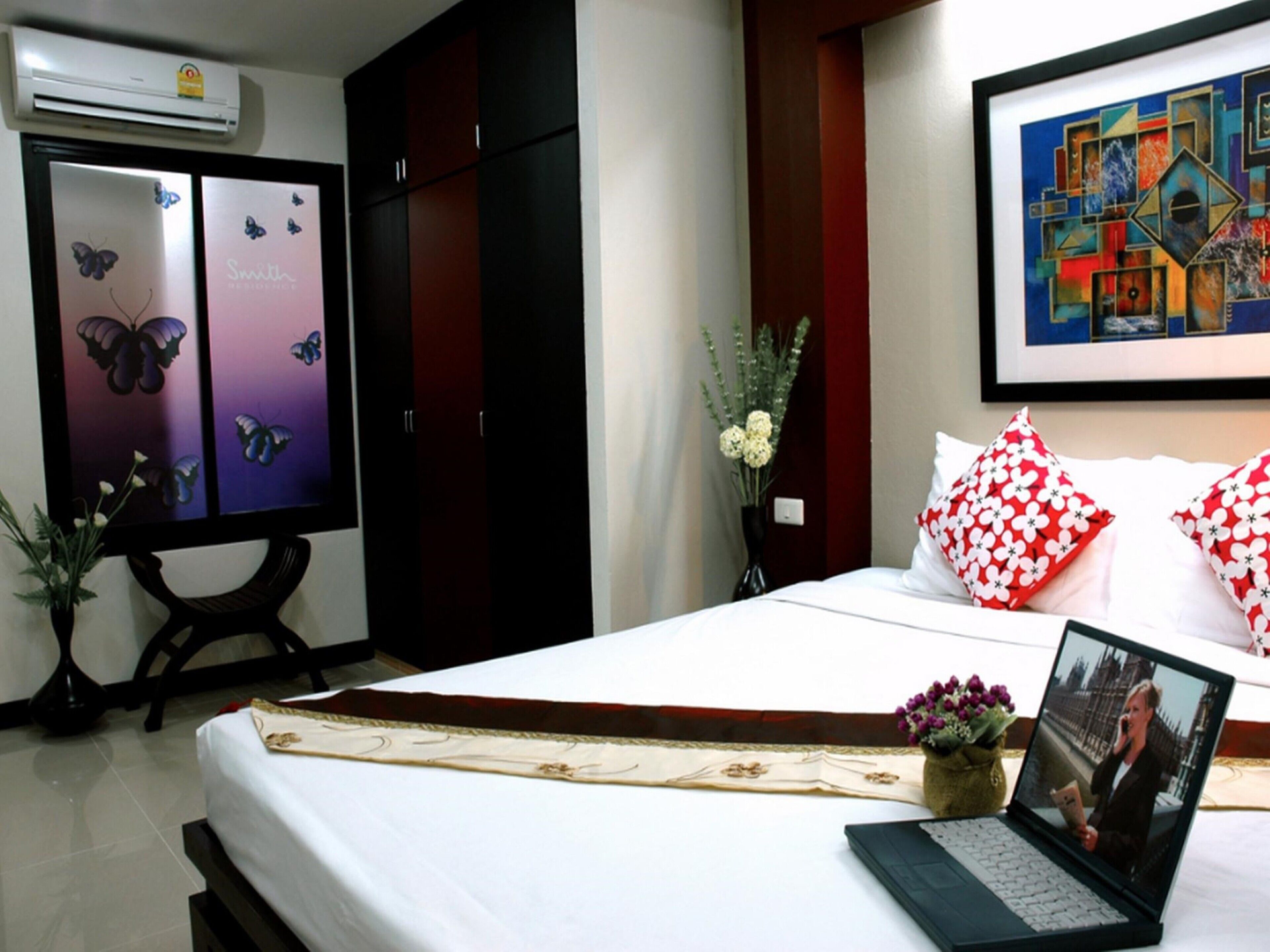 Quarto de hotel com cama, laptop, um vaso com flores, um quadro na parede e um guarda-roupa com adesivos de borboletas.