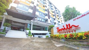 Entrada moderna de hotel com escadaria para o saguão, uma placa grande com o nome do hotel e um canteiro de flores coloridas.