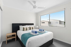 Quarto com cama, mesa de cabeceira, ventoinha no teto, janela com vista para edifícios e luminária na parede.