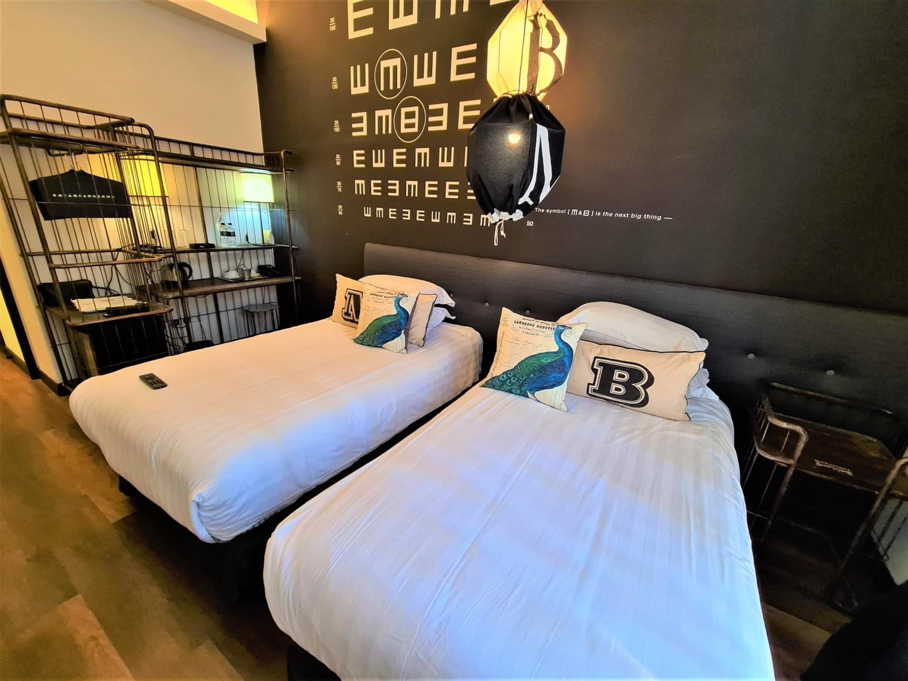Quarto de hotel com duas camas, cada uma com roupa de cama branca e almofadas decorativas, cabeceira preta e parede com um grande letreiro iluminado com a letra 'B'.