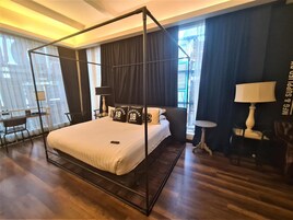 Uma cama de dossel com colcha branca, dois travesseiros com a inscrição 'Boutique 18', uma mesinha de cabeceira com luminária e vista para a cidade através de grandes janelas.