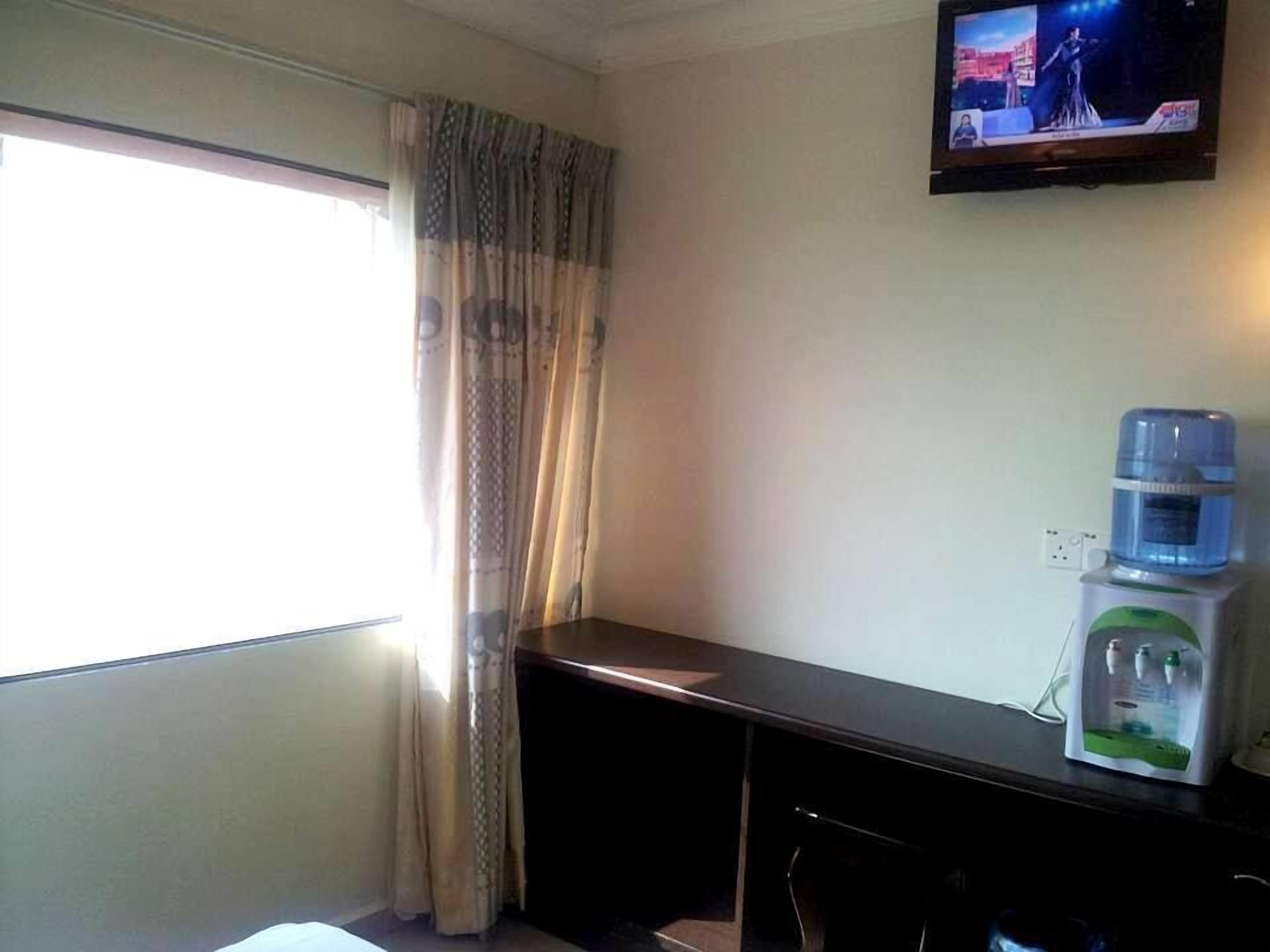 Quarto de hotel com uma janela ampla, cortina, televisão de tela plana na parede e um dispensador de água na mesa.