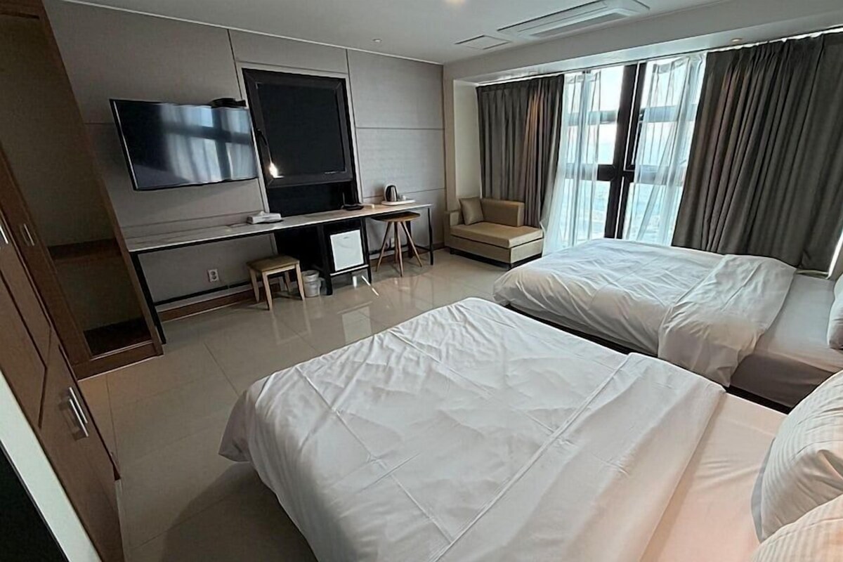 Quarto de hotel com duas camas, uma escrivaninha, uma televisão e um sofá.