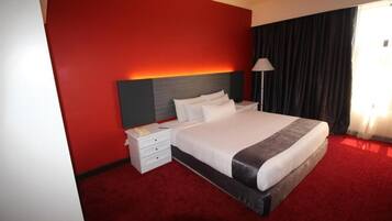 Quarto de hotel com uma cama grande, uma mesinha de cabeceira, um abajur e um tapete vermelho.