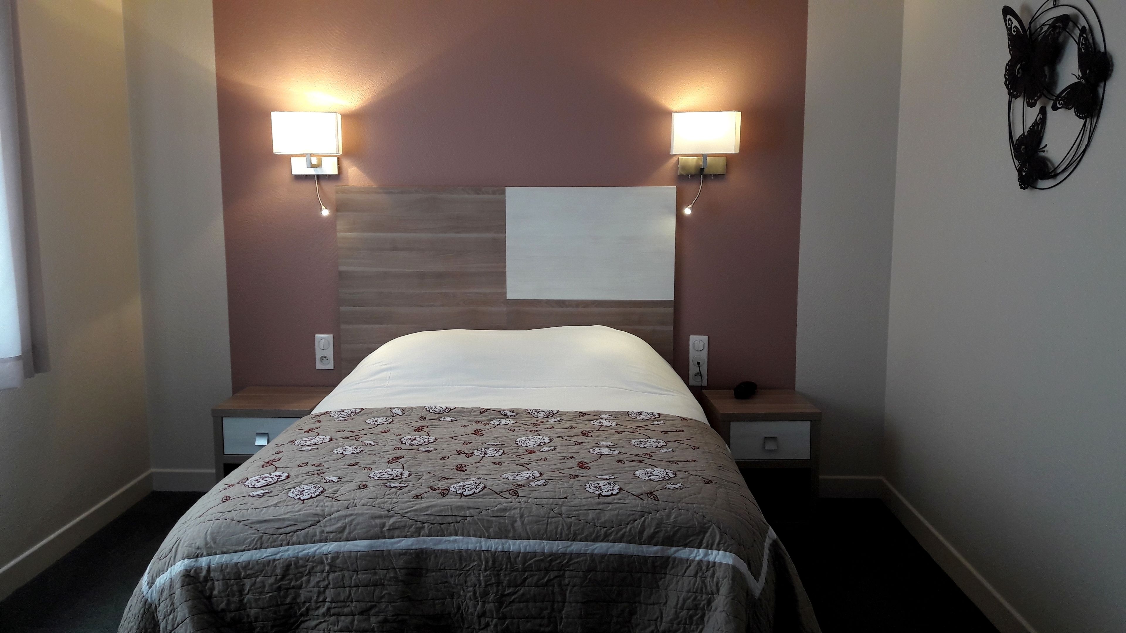 Quarto de hotel com cama, duas luminárias de apoio, mesa de cabeceira e luminária na parede.
