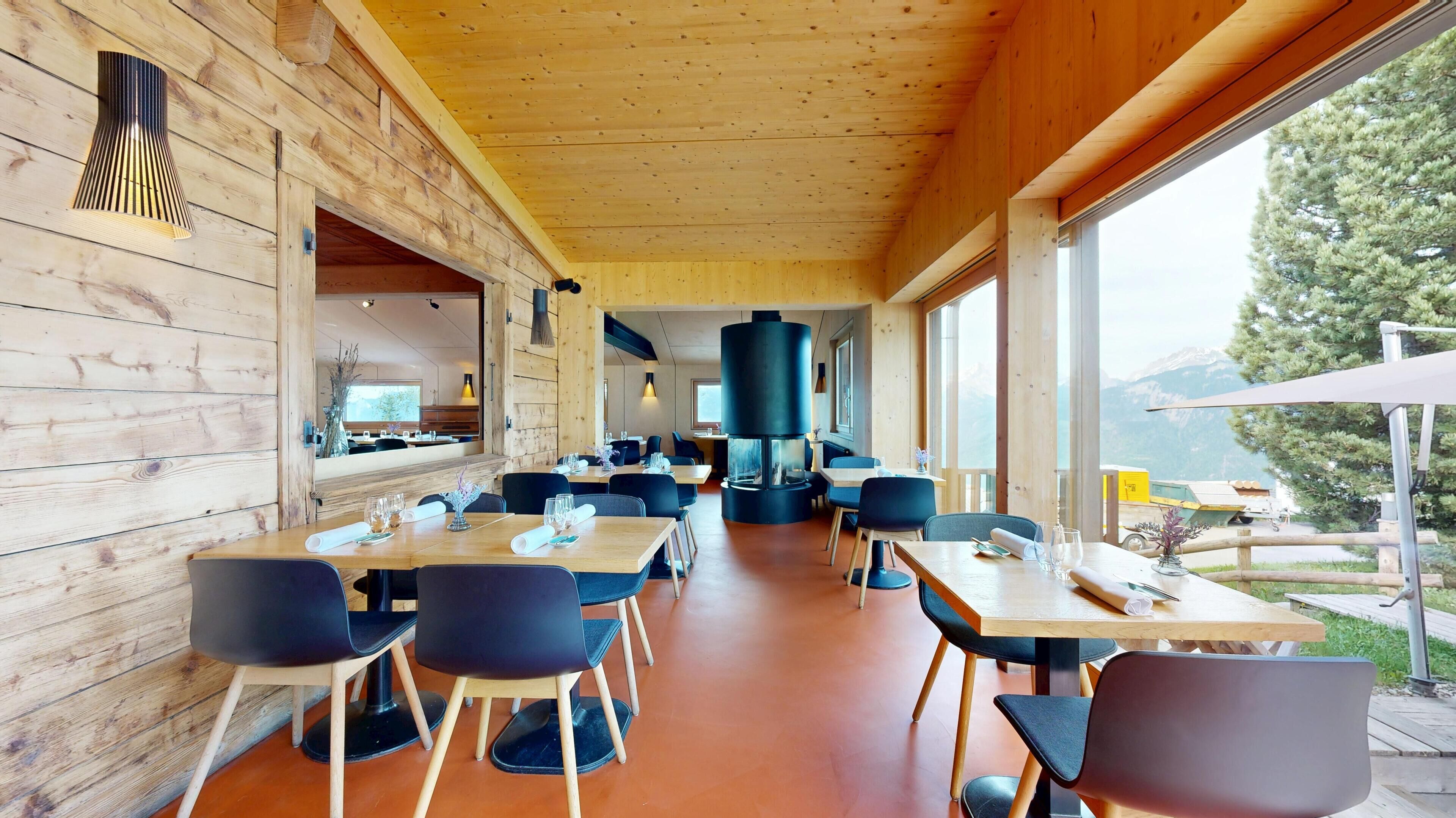 Restaurante com paredes de madeira e uma lareira central. Mesas dispostas para refeições, e grandes janelas com vista para o exterior.