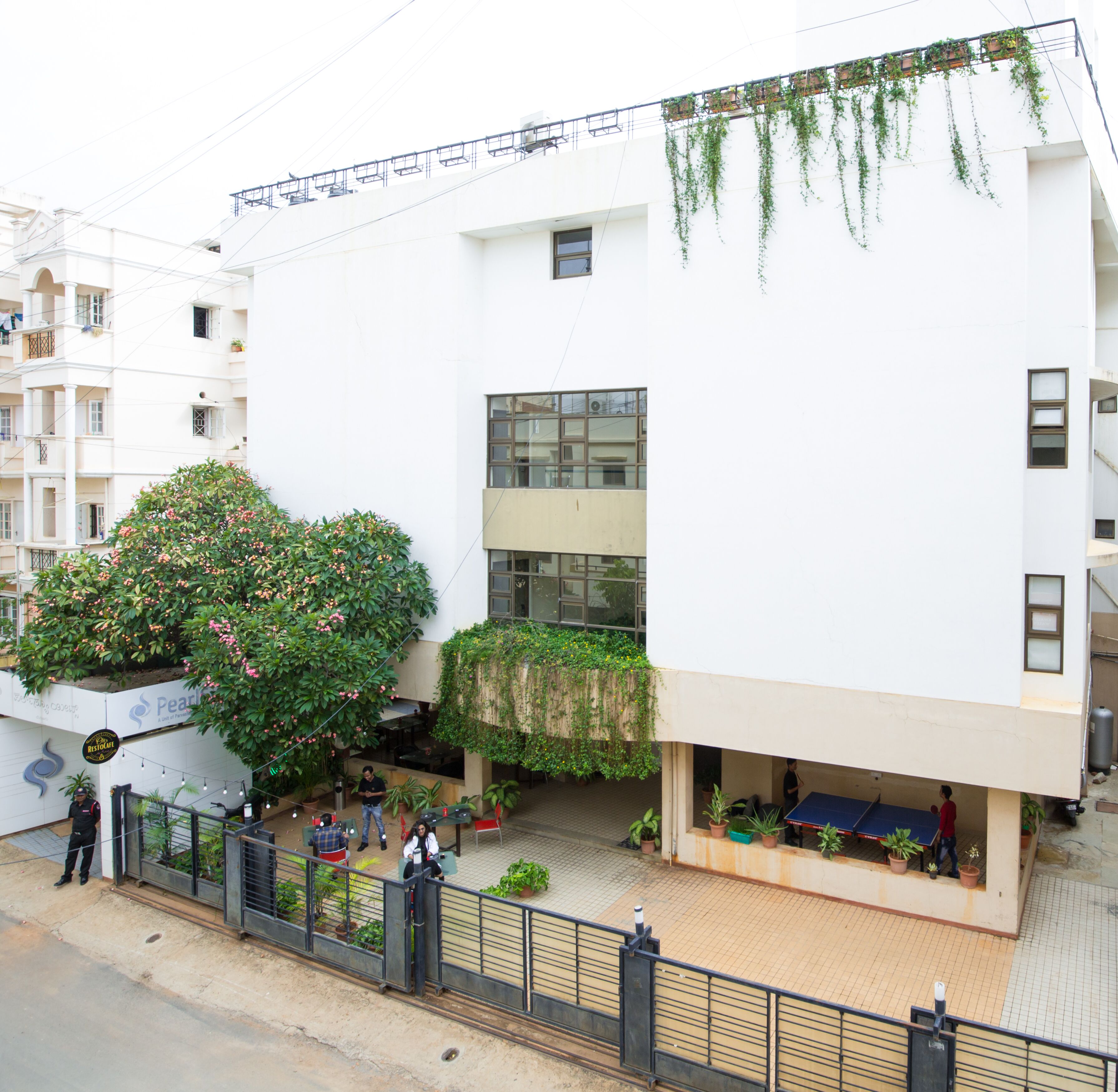 Um edifício moderno com fachada branca, uma grande árvore com flores cor-de-rosa e uma pequena área externa com mesas e cadeiras.