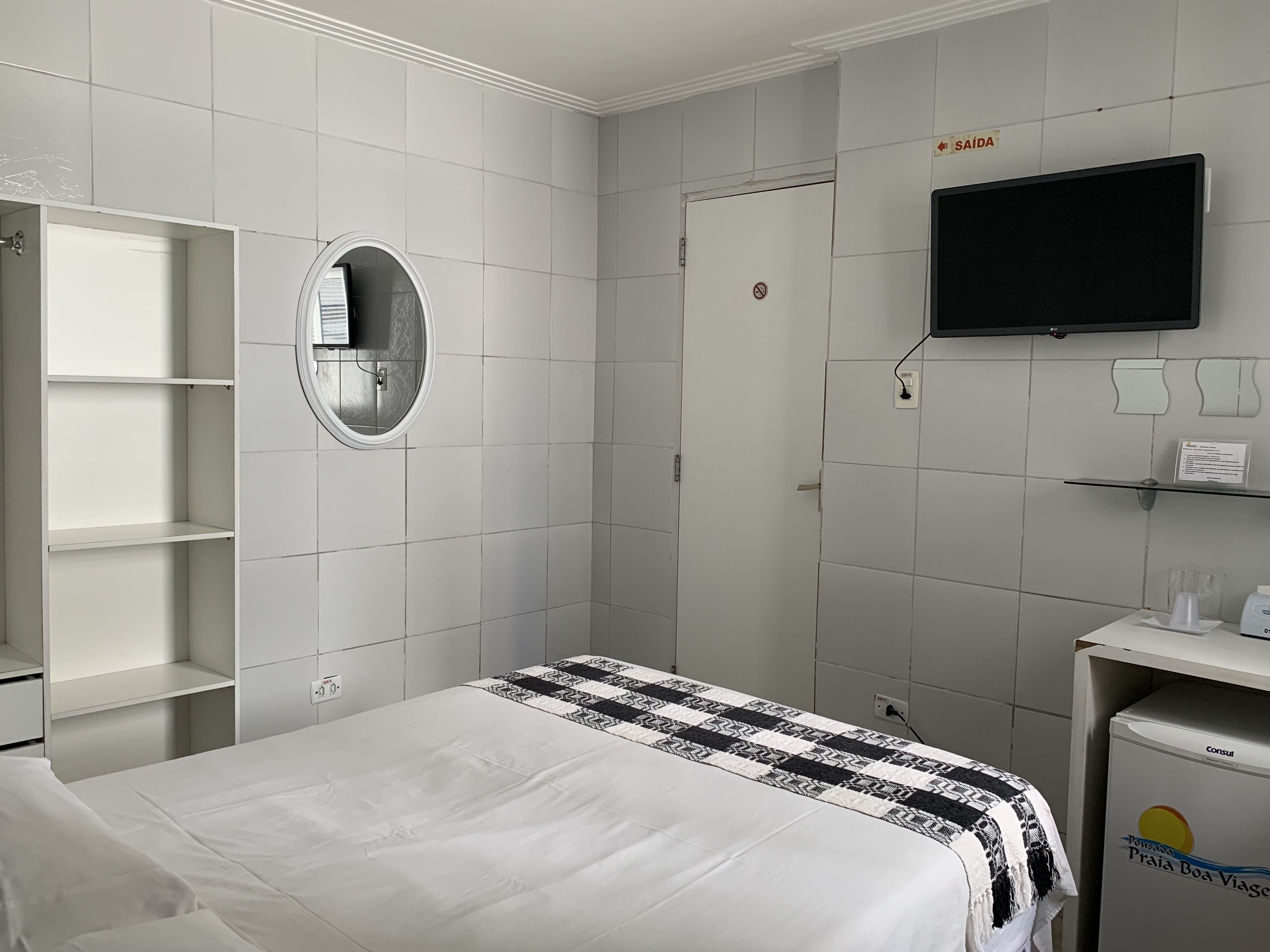 Quarto de hotel com cama, guarda-roupa, espelho redondo, televisão e minibar.