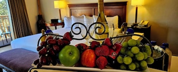 Quarto de hotel com uma cama, mesa de cabeceira, luminária e bandeja com frutas e uma garrafa.