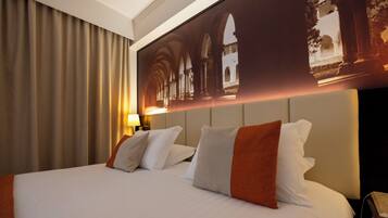 Quarto de hotel com uma cama grande, duas almofadas, cabeceira, mesa de cabeceira, abajur e um mural na parede.