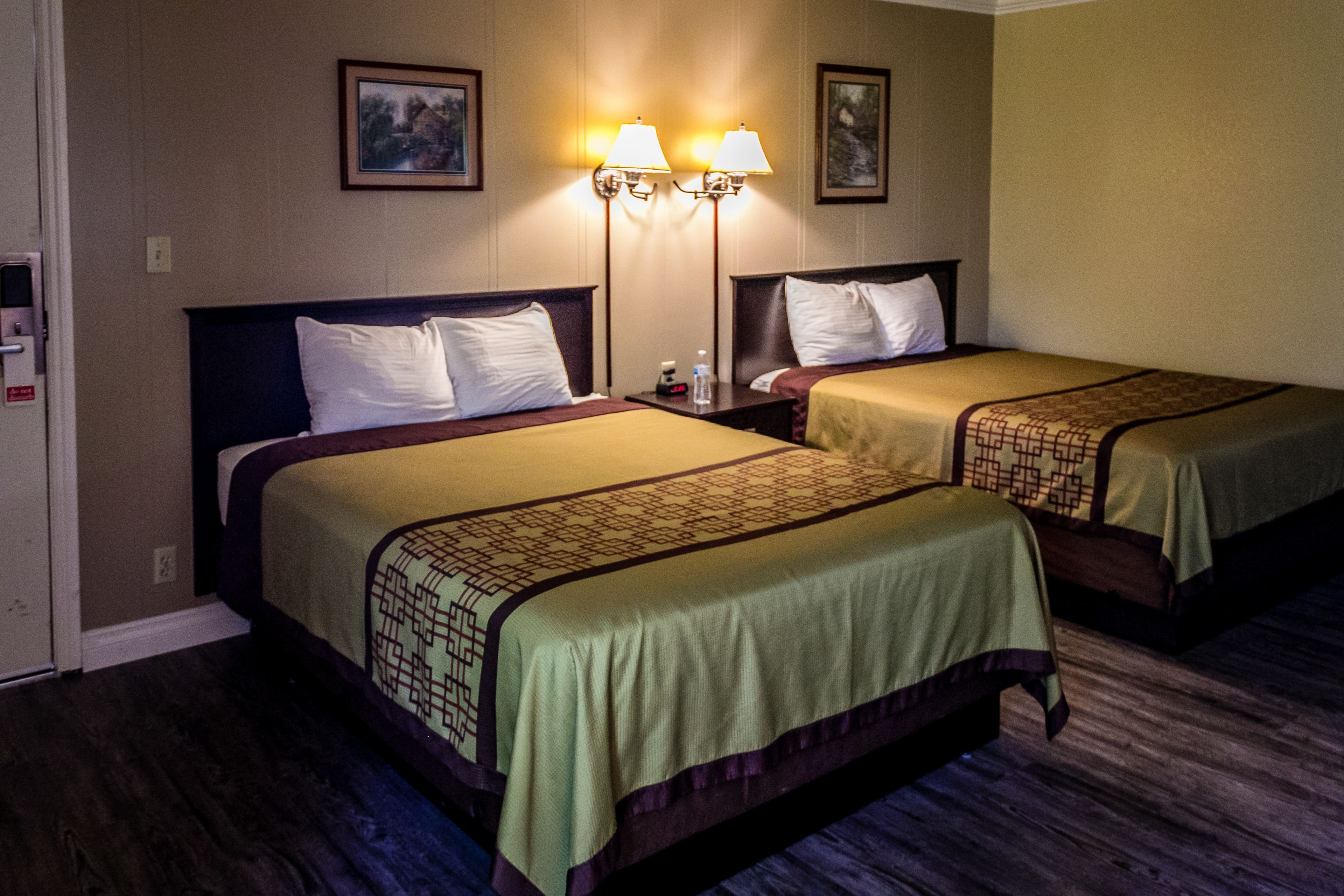 Quarto de hotel com duas camas, cada uma com colcha e almofadas estampadas, uma mesa de cabeceira com abajur e uma porta à esquerda.