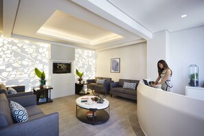 Sala de estar moderna com sofá, poltrona e mesa de centro. Há uma recepção com um recepcionista.