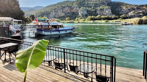 Um rio com barcos, um cais com mesas e cadeiras, e montanhas ao fundo.