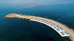 Vista aérea de um resort costeiro com uma longa e sinuosa passarela bordeada por cadeiras de praia azuis e guarda-sóis, que leva a uma península rochosa com um pequeno edifício.