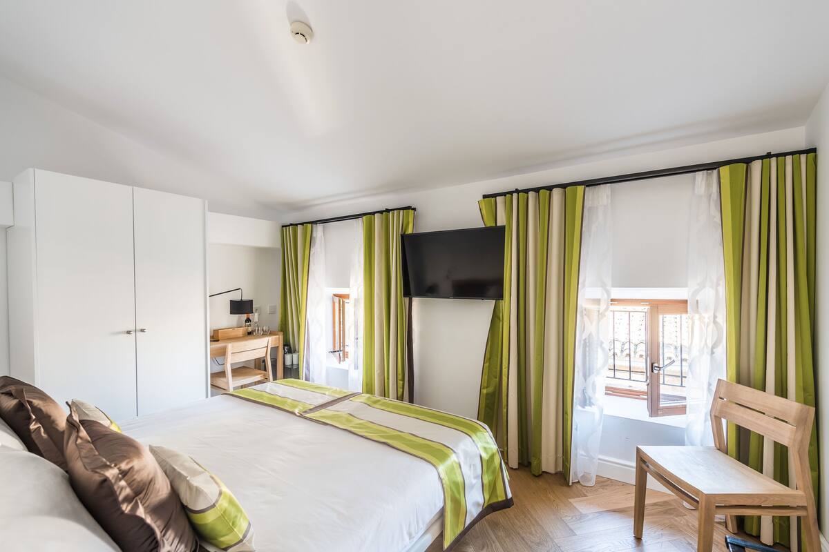 Quarto com cama, escrivaninha, cadeira e televisão na parede.