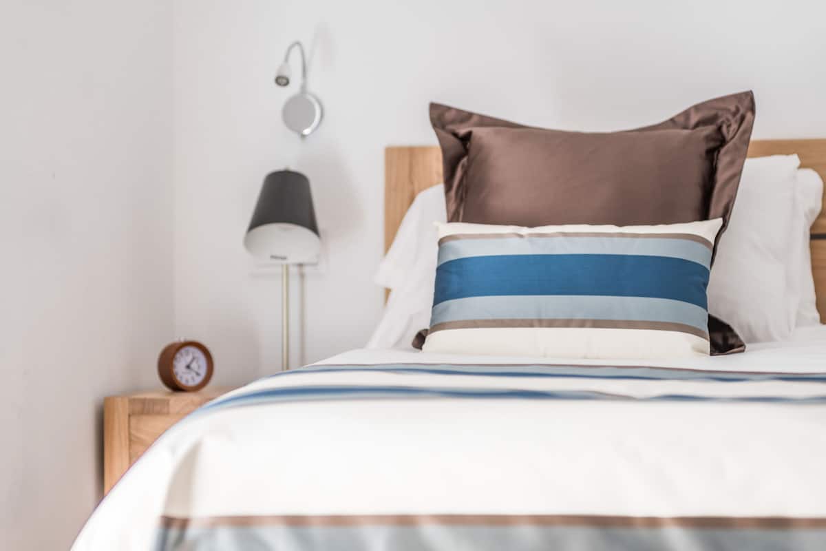 Uma cama bem arrumada com roupa de cama listrada, um abajur e um relógio em uma mesa de cabeceira de madeira.