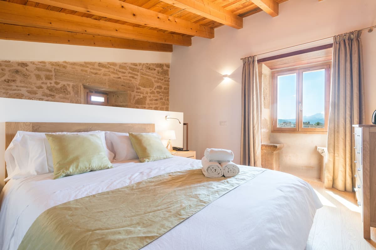 Quarto com parede de pedra, vigas de madeira, cama com lençóis brancos, mesa de cabeceira e janela com cortinas.