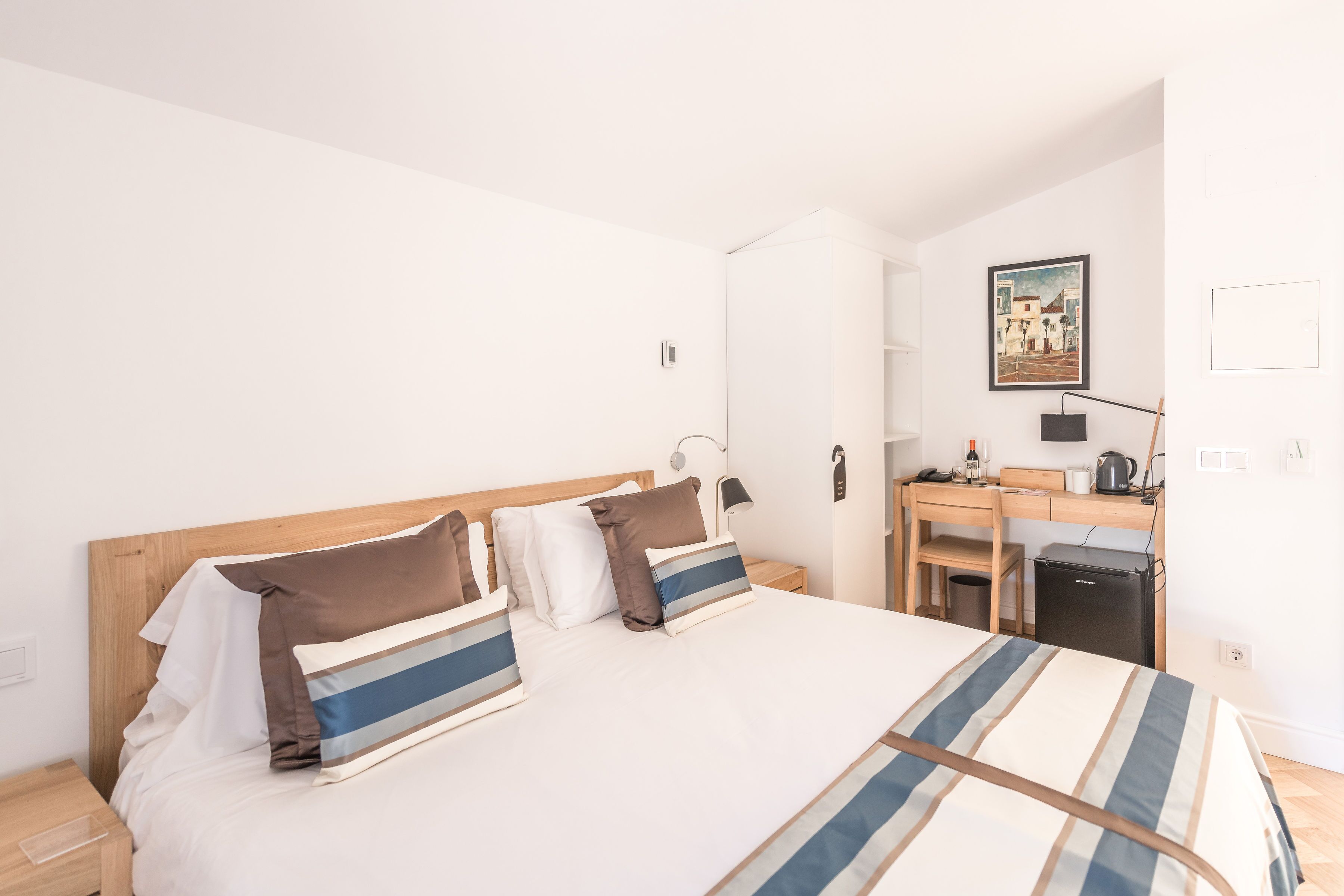 Quarto de hotel com uma cama, uma escrivaninha, uma cadeira, um mini-frigobar e um quadro na parede.