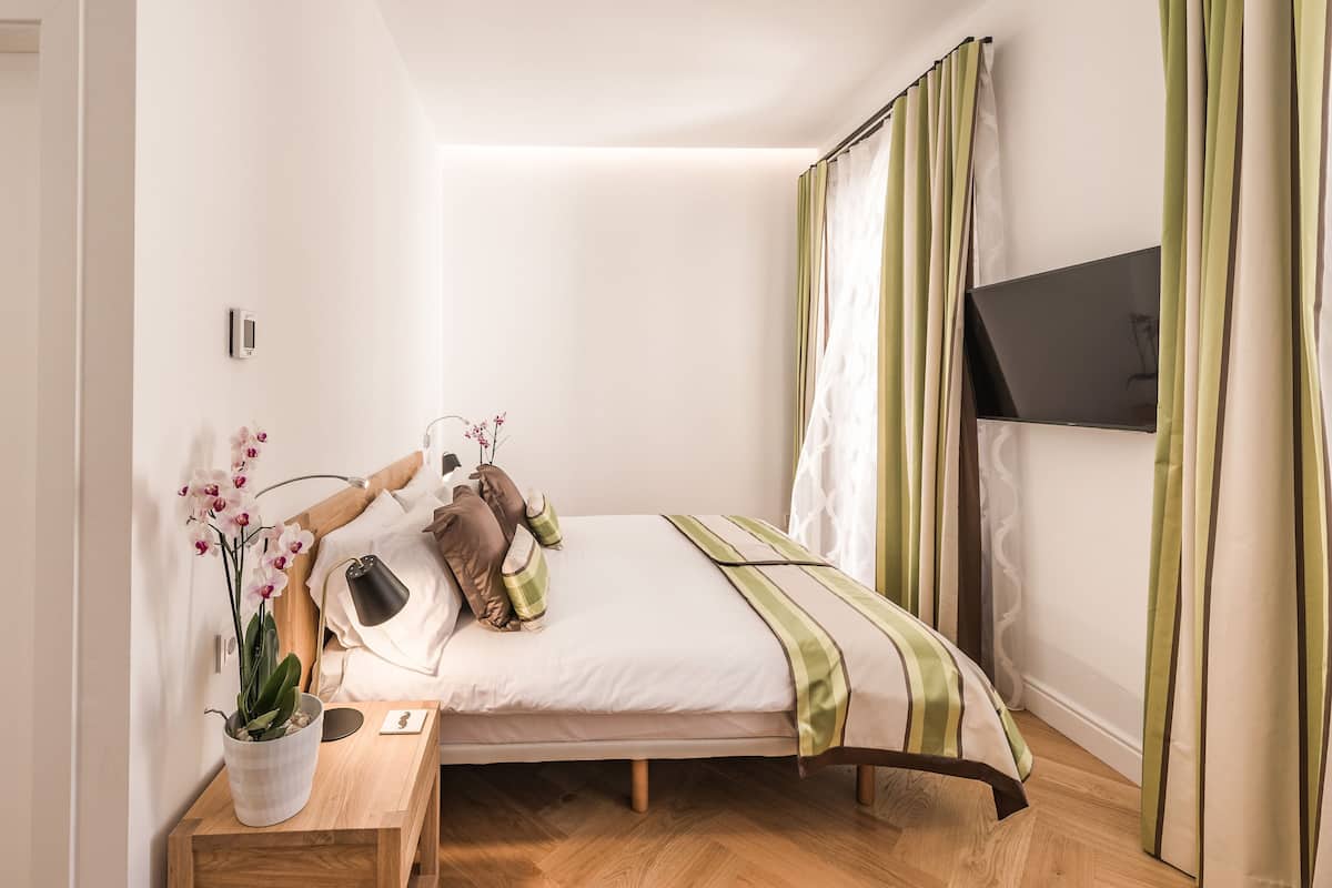Quarto arrumado com cama, mesa de cabeceira, abajur e uma orquídea em vaso.