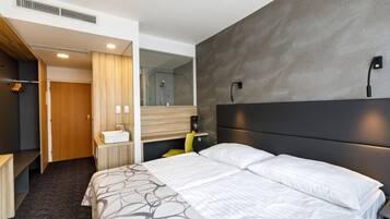 Quarto de hotel moderno com cama grande, mesa de cabeceira, espelho e banheiro visível através de uma porta aberta.