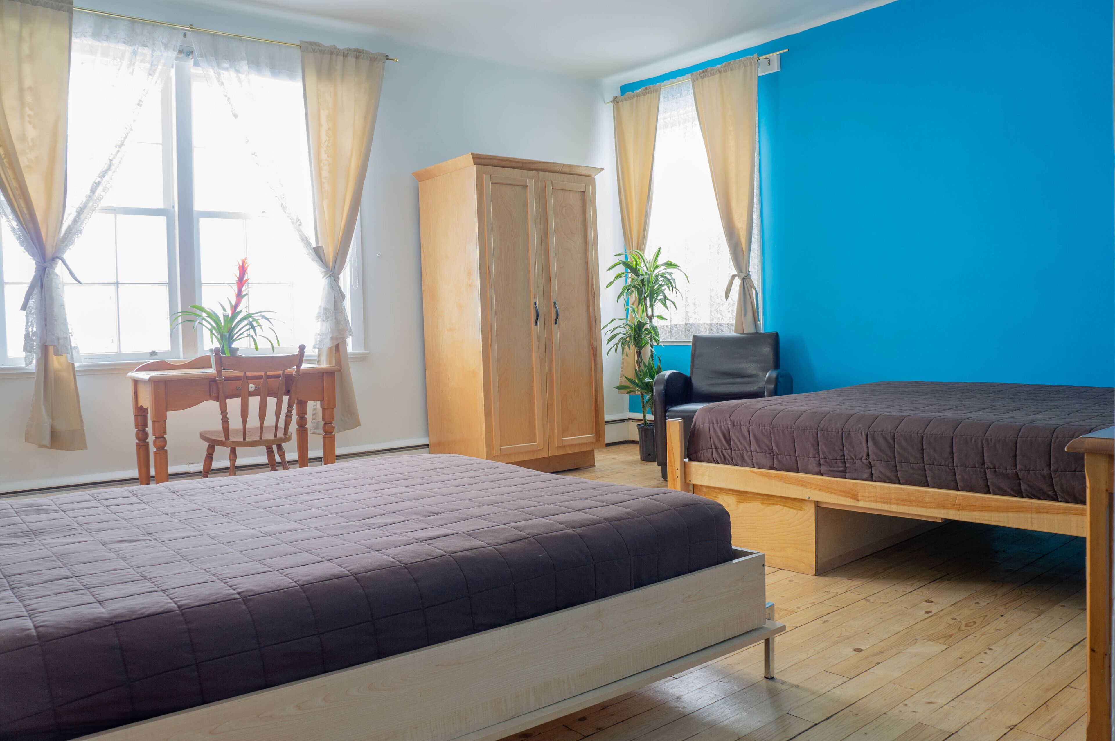 Quarto com duas camas, um guarda-roupa de madeira, uma escrivaninha com cadeira e um vaso de planta.