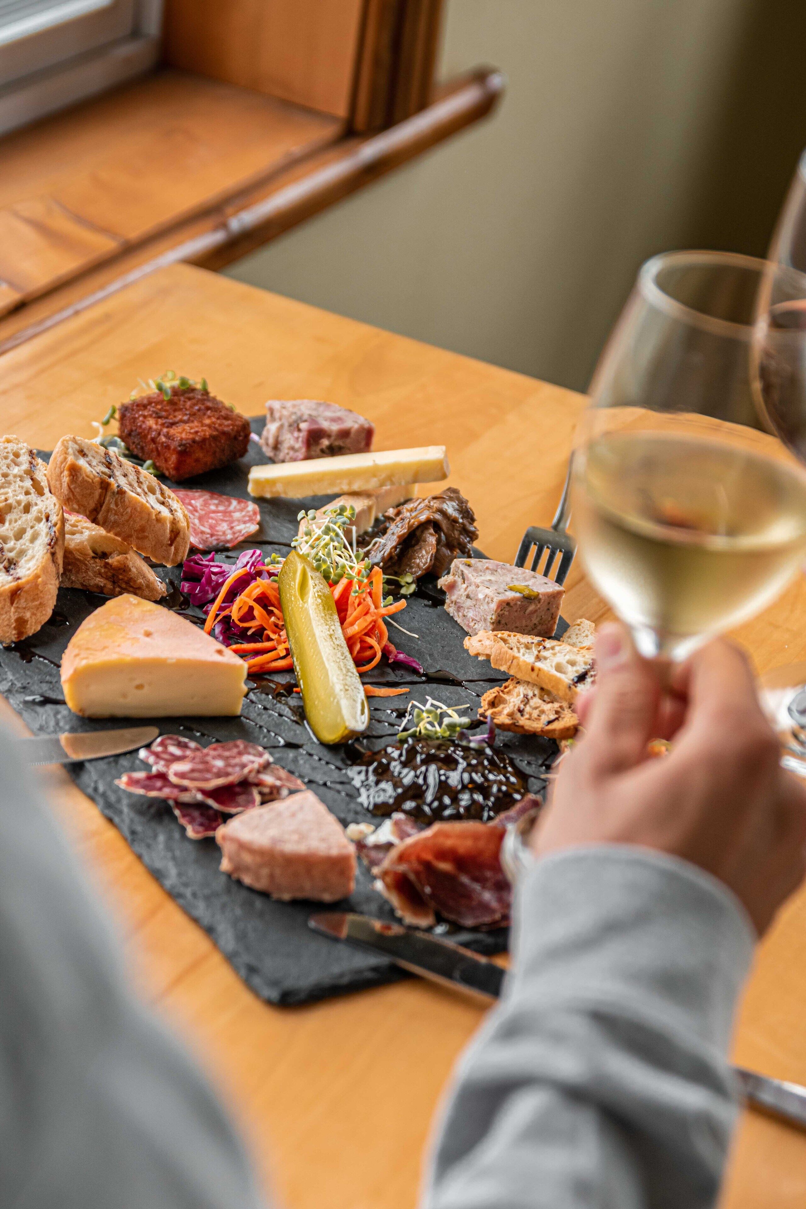 Uma mesa com diversos queijos, carnes e vegetais, e uma pessoa segurando um copo de vinho branco.