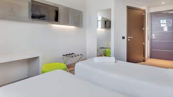 Quarto de hotel com duas camas, uma televisão fixada na parede, uma porta com puxador e uma pequena mesa com uma cadeira.