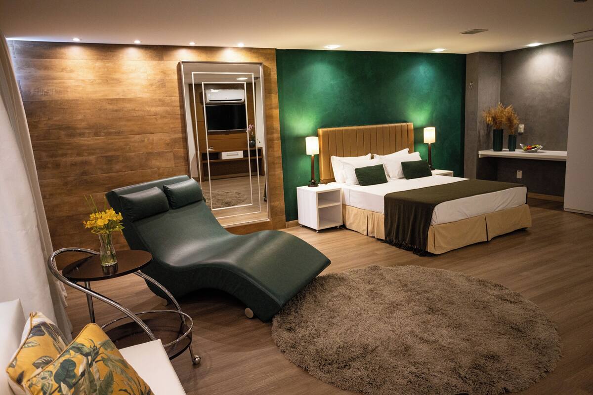 Quarto moderno com cama, espreguiçadeira, mesa pequena com um vaso e um tapete grande e fofo.