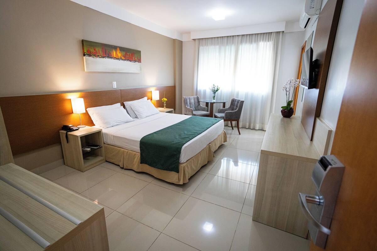 Quarto de hotel com cama, mesas de cabeceira, escrivaninha, cadeiras e um quadro na parede.