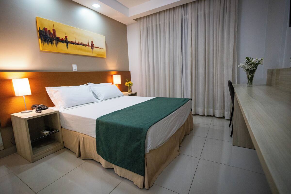 Quarto de hotel com cama, luminárias de cabeceira, mesa de cabeceira, escrivaninha e uma cadeira.