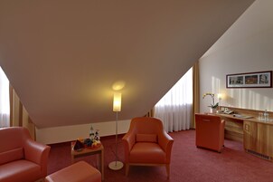 Quarto de hotel com teto inclinado, uma escrivaninha com telefone, uma cadeira, uma mesinha com frutas e um abajur.