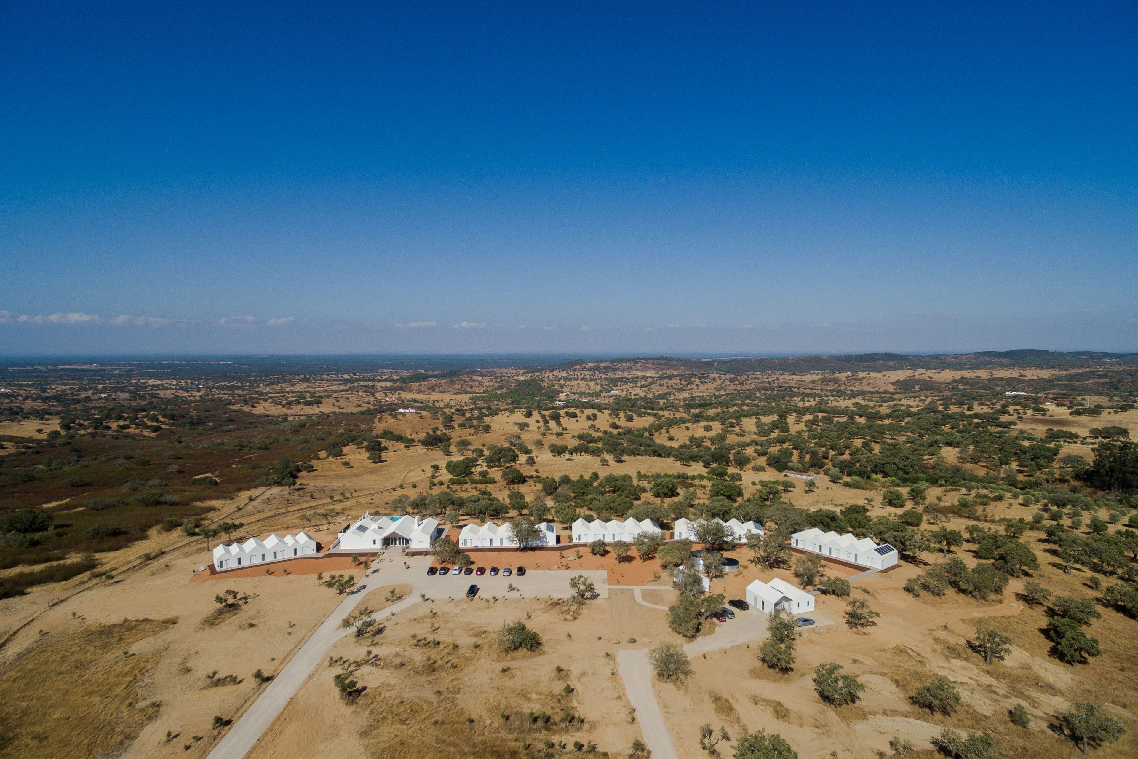 Vista aérea de um resort com vários edifícios brancos dispostos em formato de semicírculo, cercado por terreno seco e gramado, e vegetação esparsa.