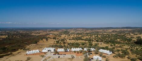 Vista aérea de um resort com vários edifícios brancos dispostos em formato de semicírculo, cercado por terreno seco e gramado, e vegetação esparsa.