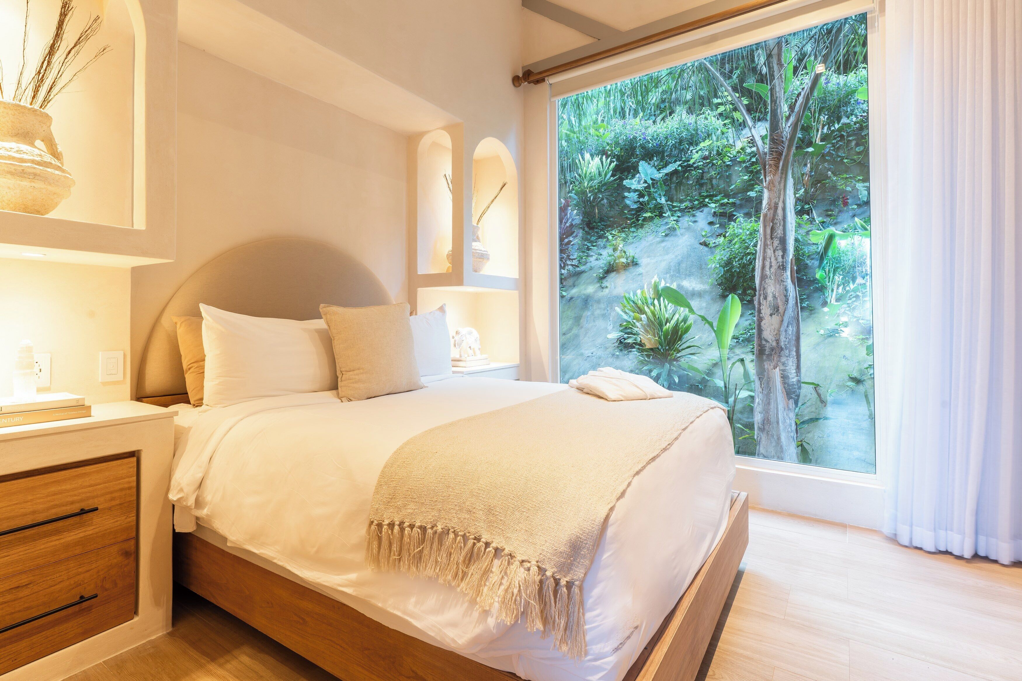 Quarto com uma grande janela, cama com roupa de cama branca, mesa de cabeceira com abajur e vista para o verde lá fora.