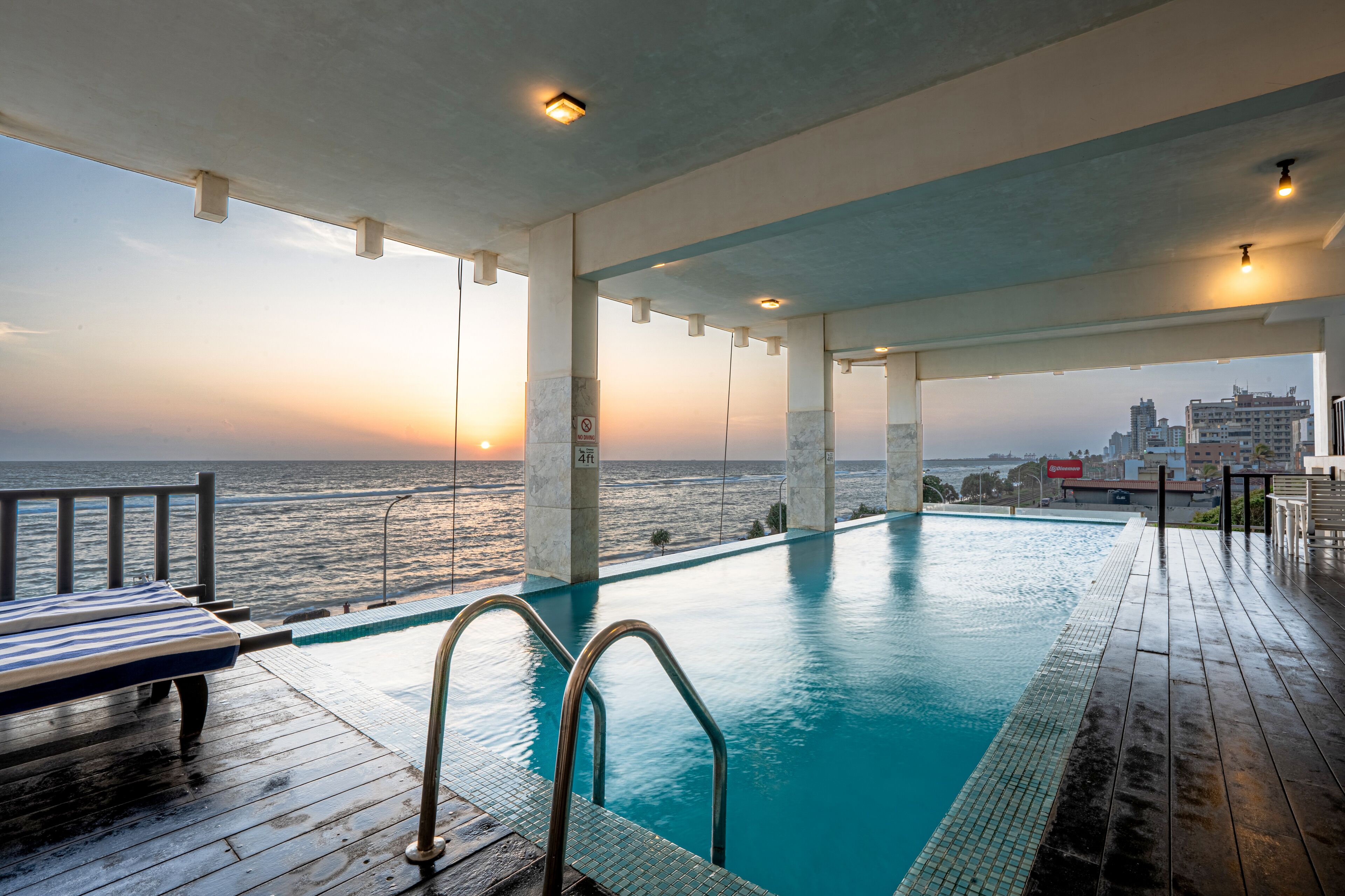 Piscina infinita com vista para o oceano ao pôr do sol, deck de madeira e área de estar com um banco.