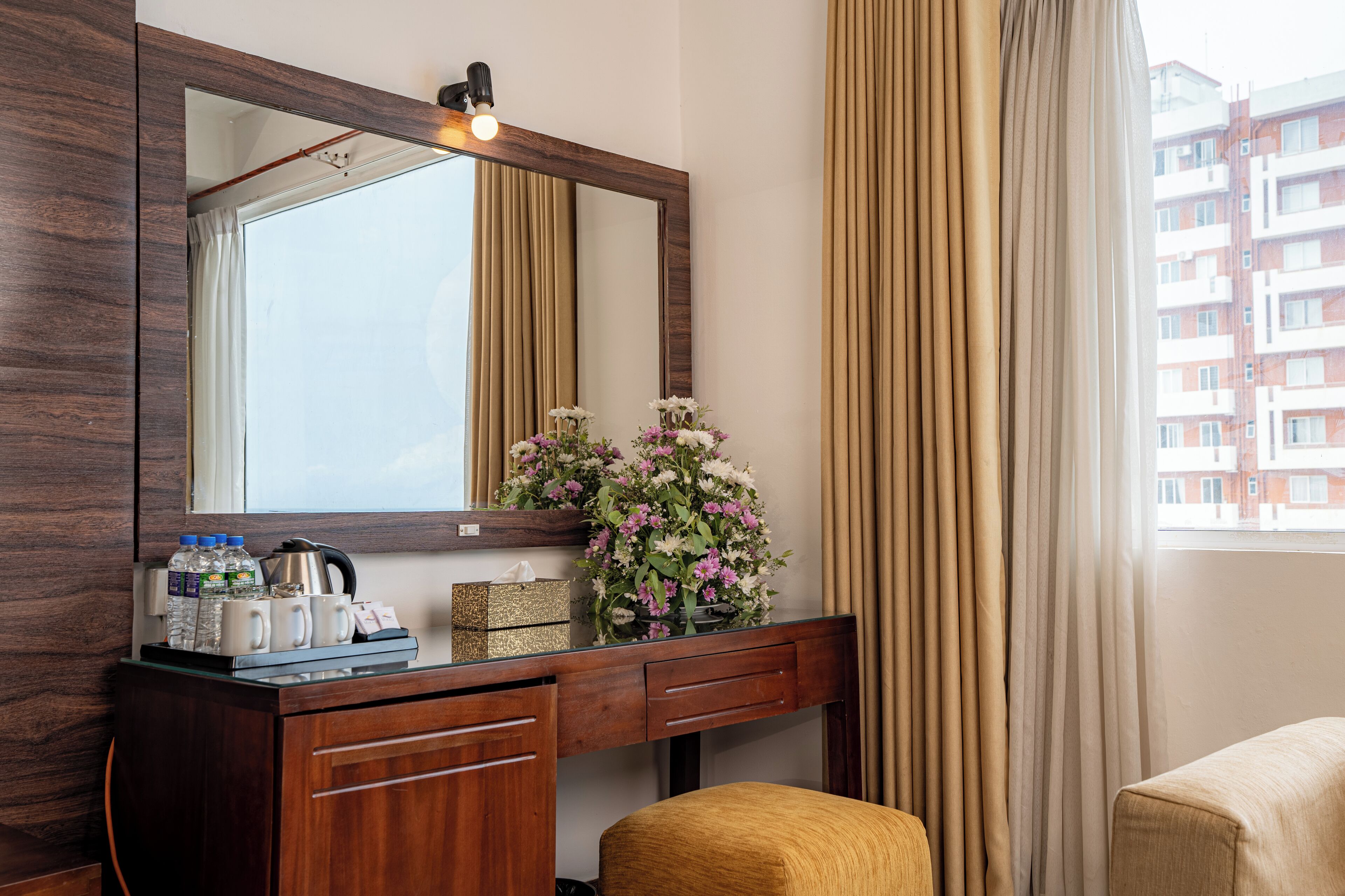 Quarto de hotel com escrivaninha de madeira, espelho, um vaso de flores e janela com cortinas.