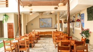 Uma ampla sala de jantar com mesas e cadeiras de madeira dispostas em fileiras, teto alto com vigas de madeira e luminárias esféricas penduradas.