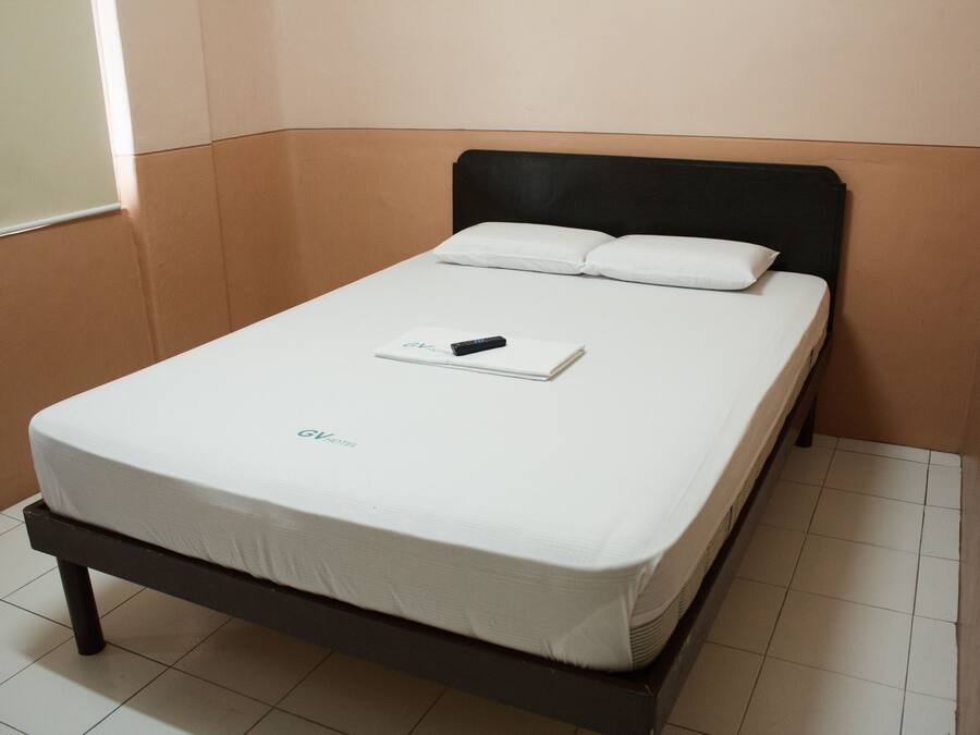 Uma cama de solteiro com cabeceira preta, roupa de cama branca e um controle remoto sobre o colchão.
