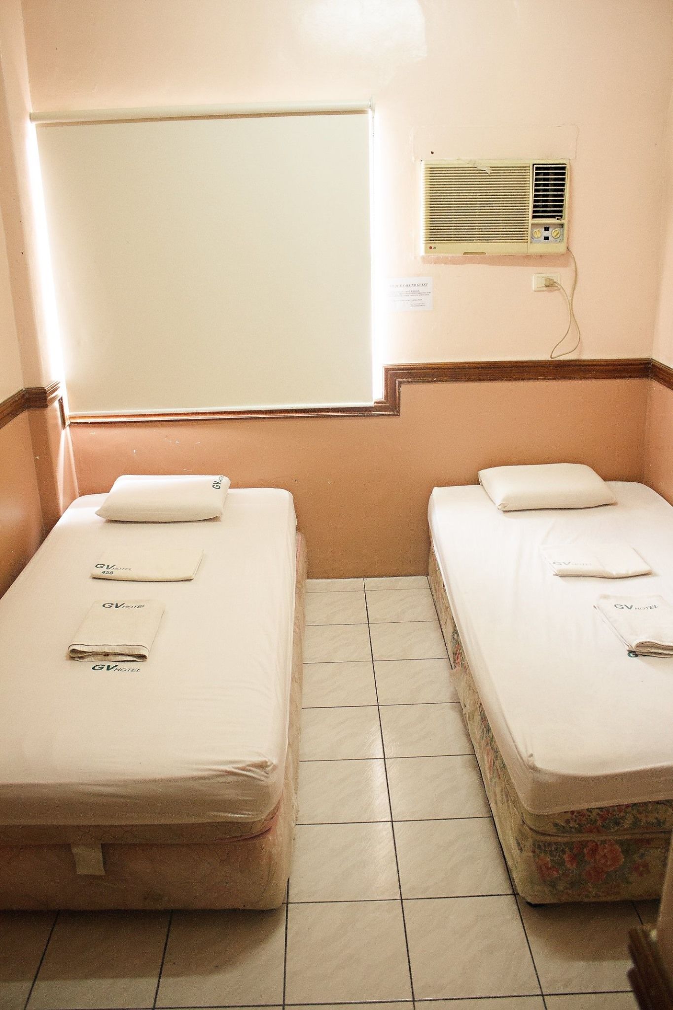 Quarto de hotel pequeno com duas camas de solteiro, uma janela, ar condicionado fixado na parede e piso de azulejos.