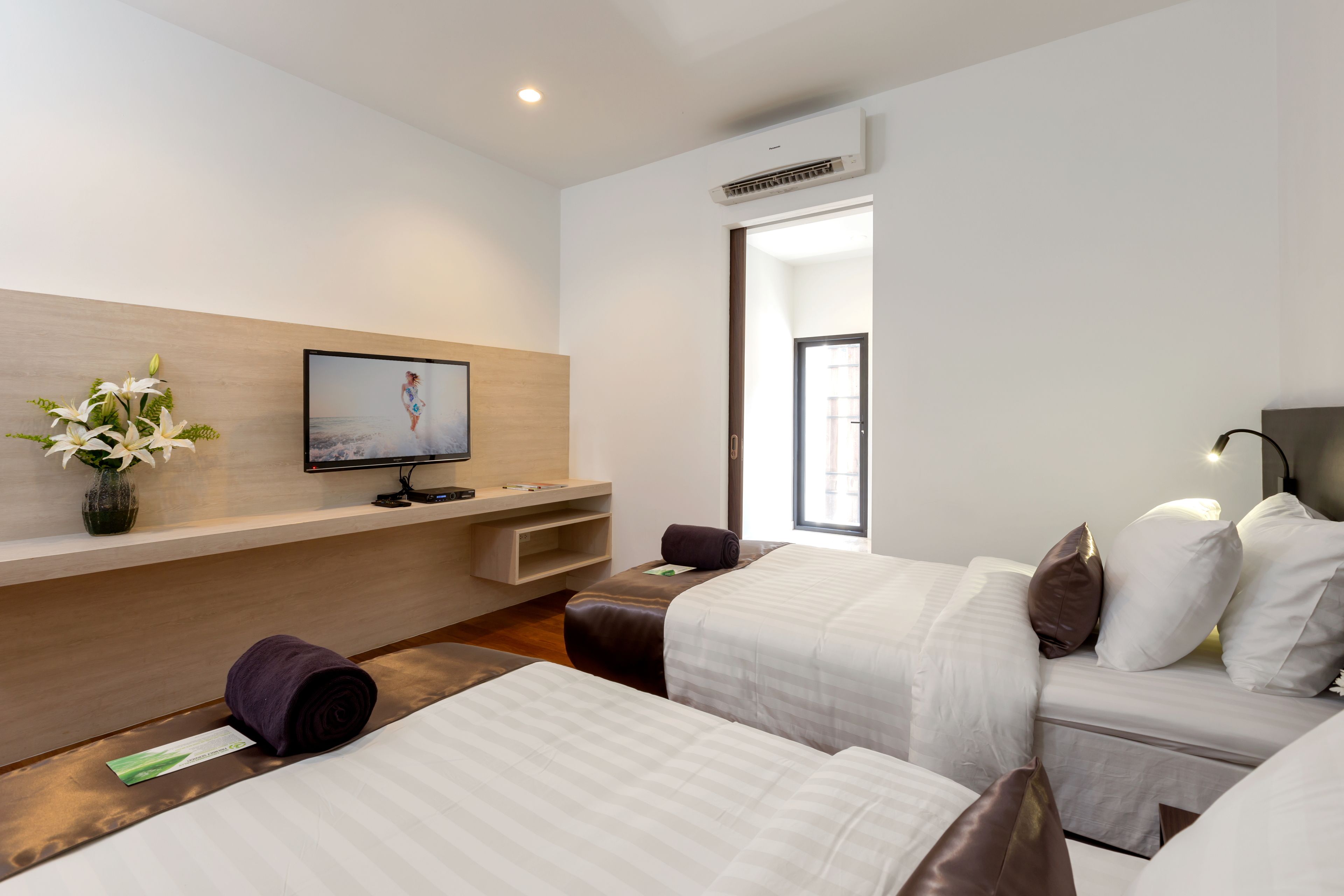 Quarto de hotel com duas camas, uma televisão em uma estante de madeira, um vaso com flores e uma janela que proporciona iluminação natural.