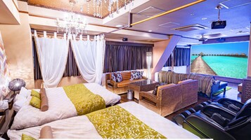 Quarto luxuoso com cama, sofá, poltronas de massagem e uma tela grande exibindo uma vista de praia.