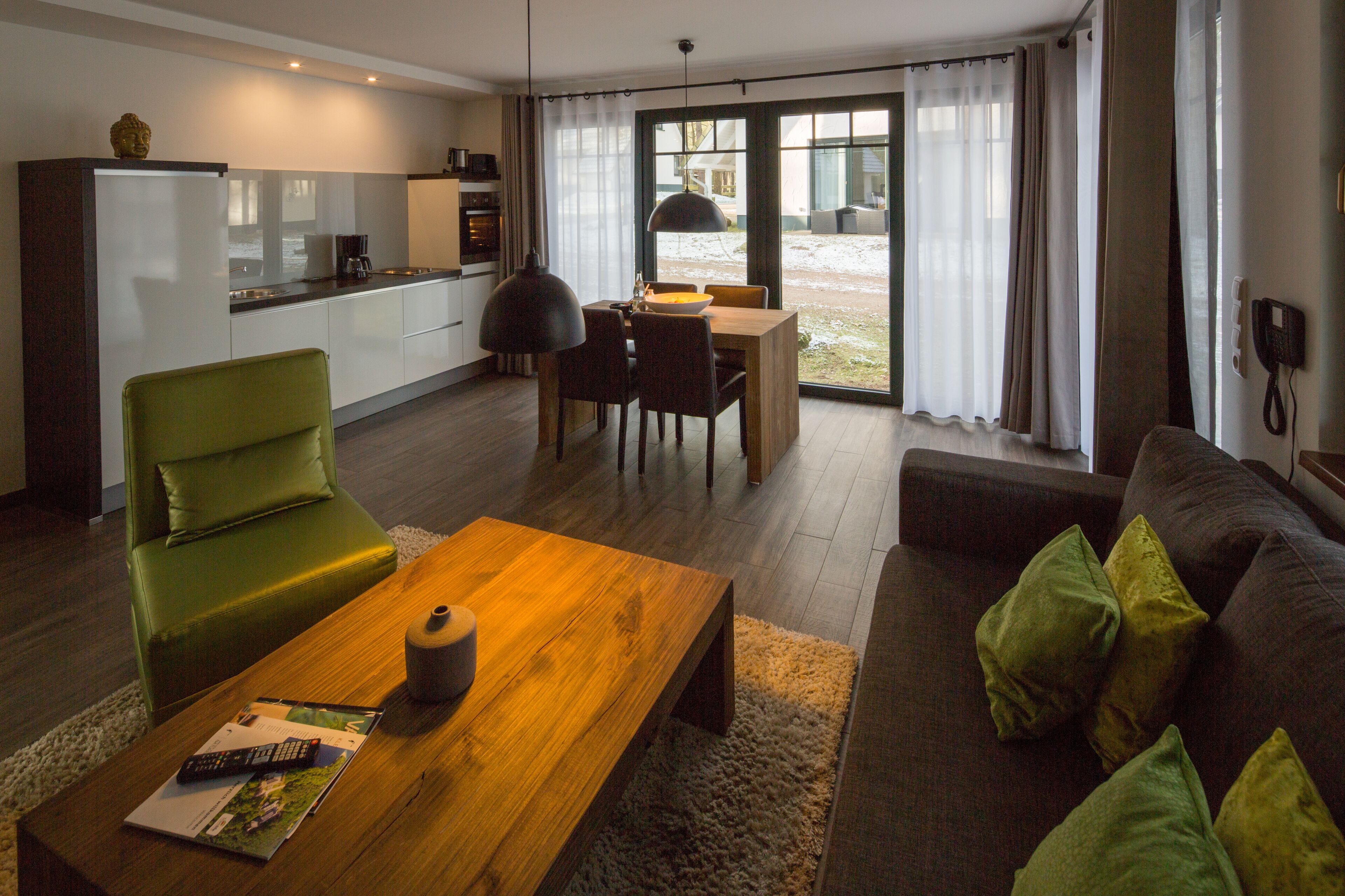 Uma sala de estar moderna com uma mesa de centro de madeira, uma poltrona verde, um sofá escuro com almofadas verdes, e um espaço de jantar com mesa e cadeiras.