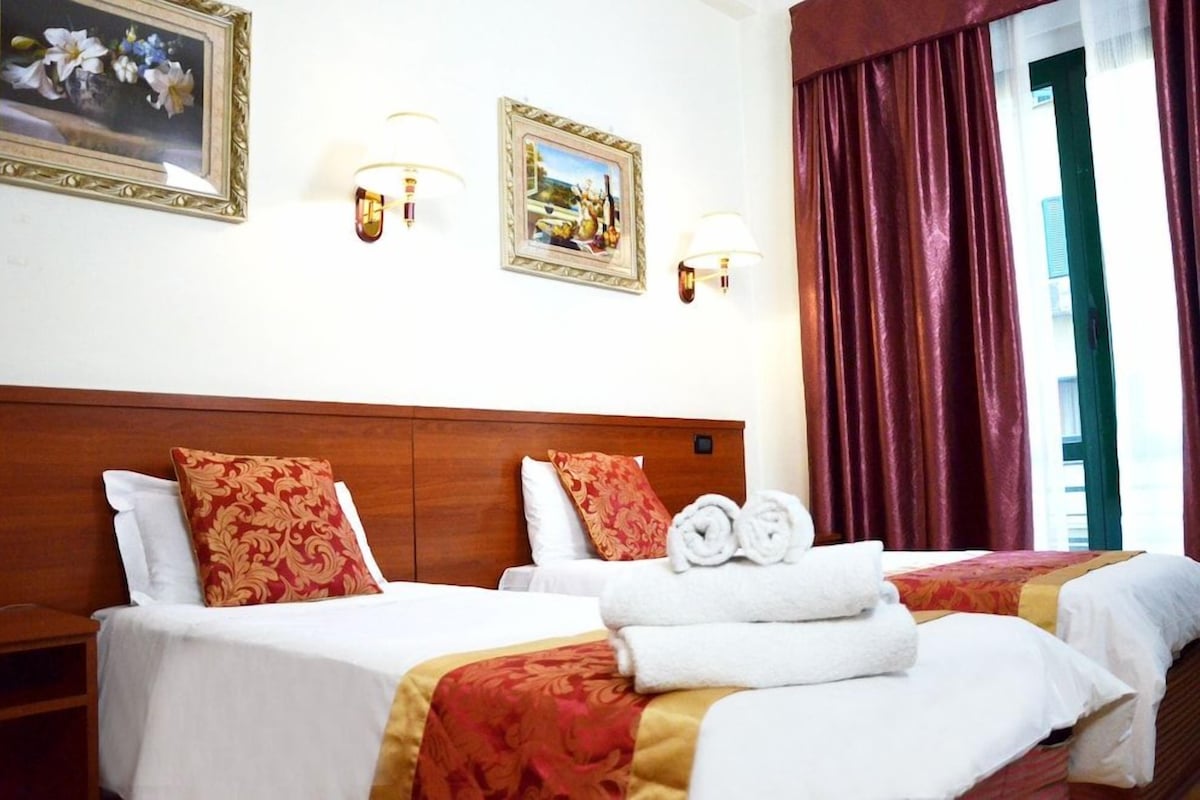 Quarto de hotel com duas camas, cabeceira de madeira, luminárias fixadas na parede e uma janela com cortinas.