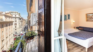 Uma varanda com vista para uma rua da cidade, uma cama com colchão azul e uma mesa pequena com abajur.